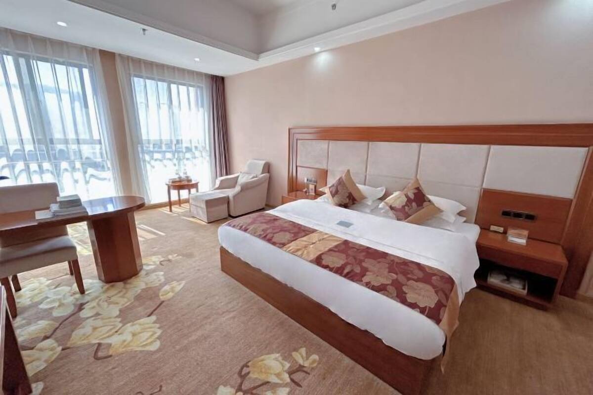 Quarto de hotel com uma cama grande, uma escrivaninha, uma cadeira e vista para o exterior através de amplas janelas.