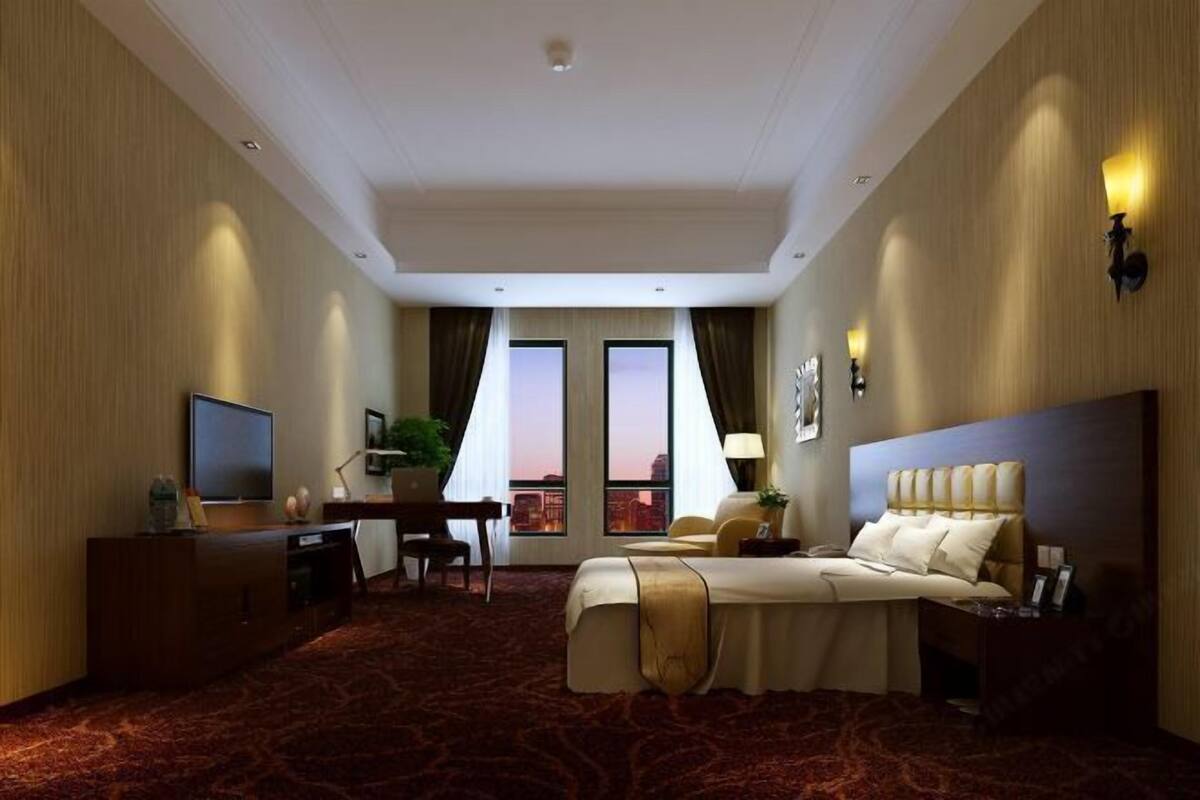 Quarto de hotel com cama, escrivaninha, televisão e vista da cidade por grandes janelas.