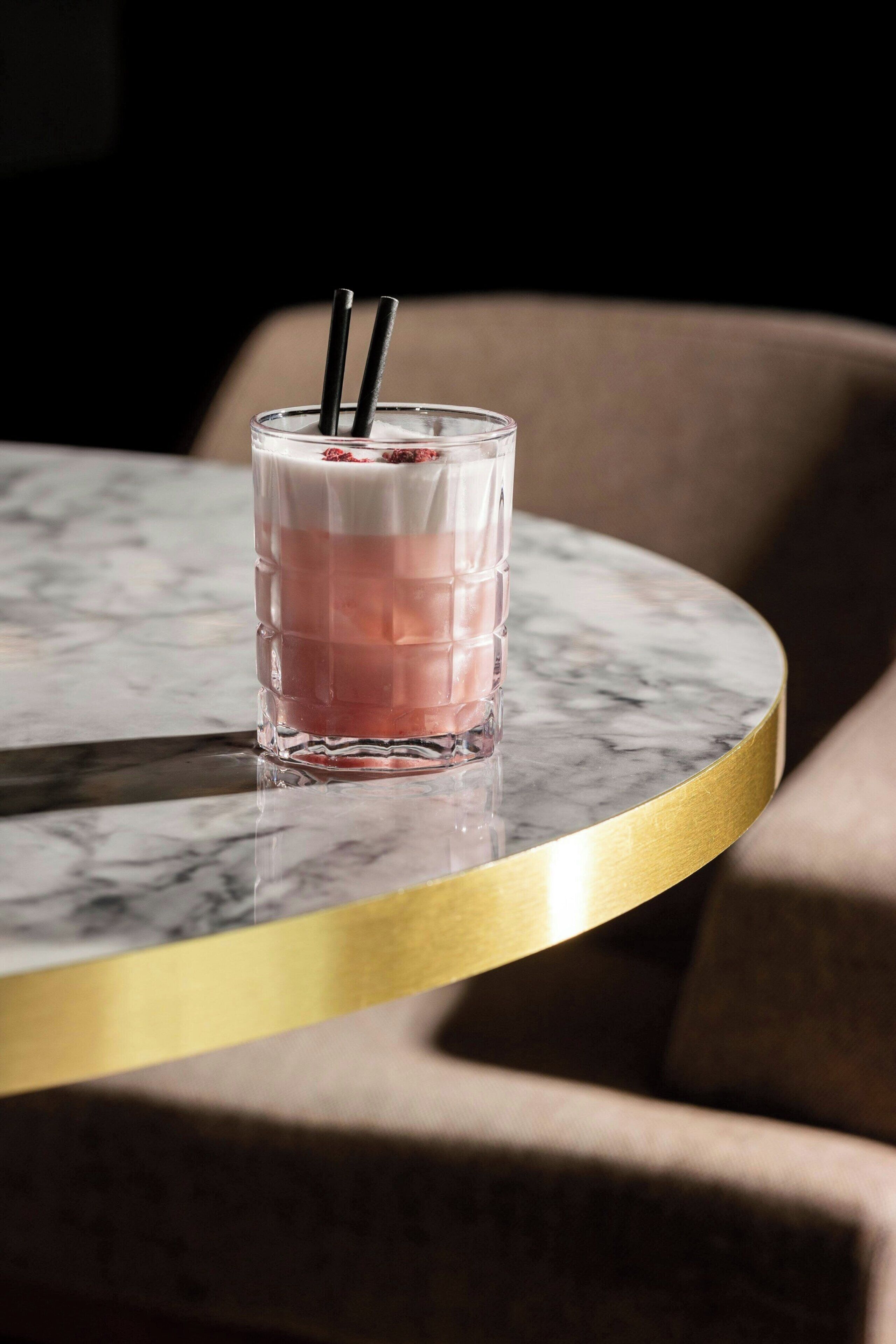 Um copo de bebida rosa com canudos pretos sobre uma mesa de mármore com borda dourada.