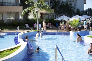 Área de piscina com espreguiçadeiras e guarda-sóis, rodeada por palmeiras e um edifício com varandas.