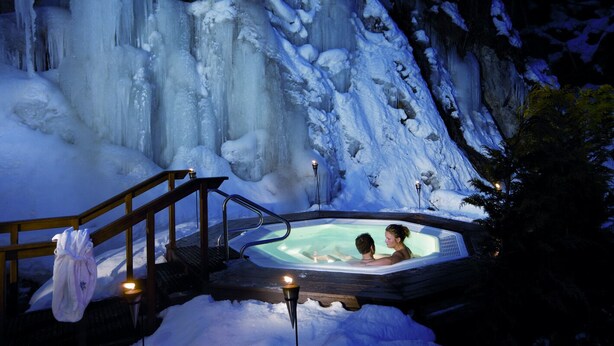 Uma banheira de hidromassagem com duas pessoas, rodeada por neve e formações de gelo, com um deck e guarda-corpos de madeira.