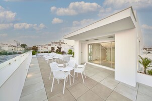Um terraço no telhado com cadeiras e mesas brancas, uma porta de vidro e vista para a paisagem urbana.