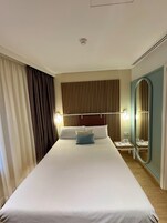 Quarto de hotel com uma cama grande, cabeceira com ripas verticais, mesa de cabeceira com luminária, espelho e piso de madeira.
