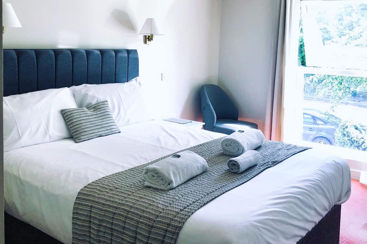 Cama bem arrumada com lençóis brancos e manta estampada, cabeceira estofada azul e luminária de cabeceira.
