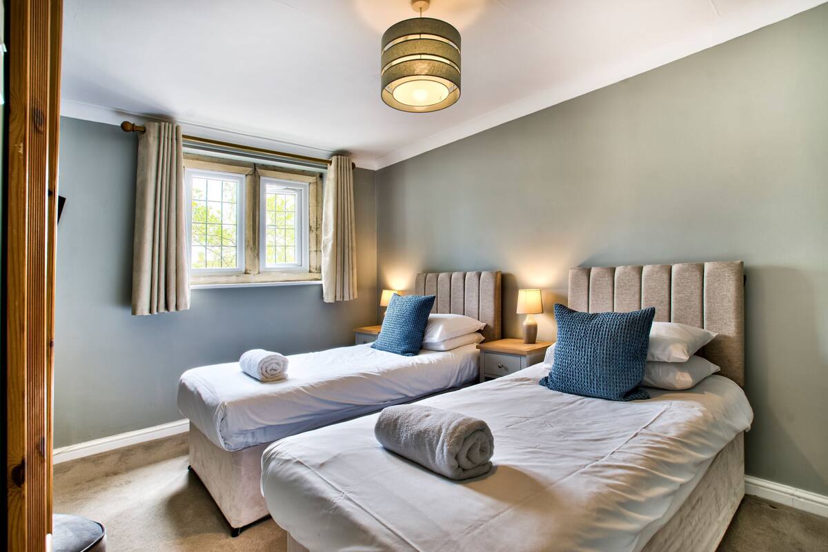 Quarto de hotel com duas camas, uma janela, uma luminária no teto e uma mesa de cabeceira com abajur.