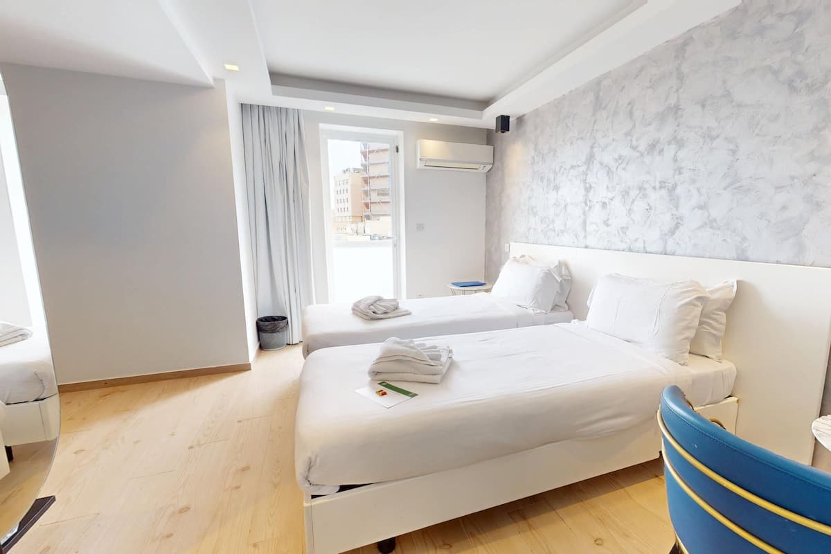Quarto de hotel com duas camas, uma mesinha, uma cadeira e vista para o exterior.