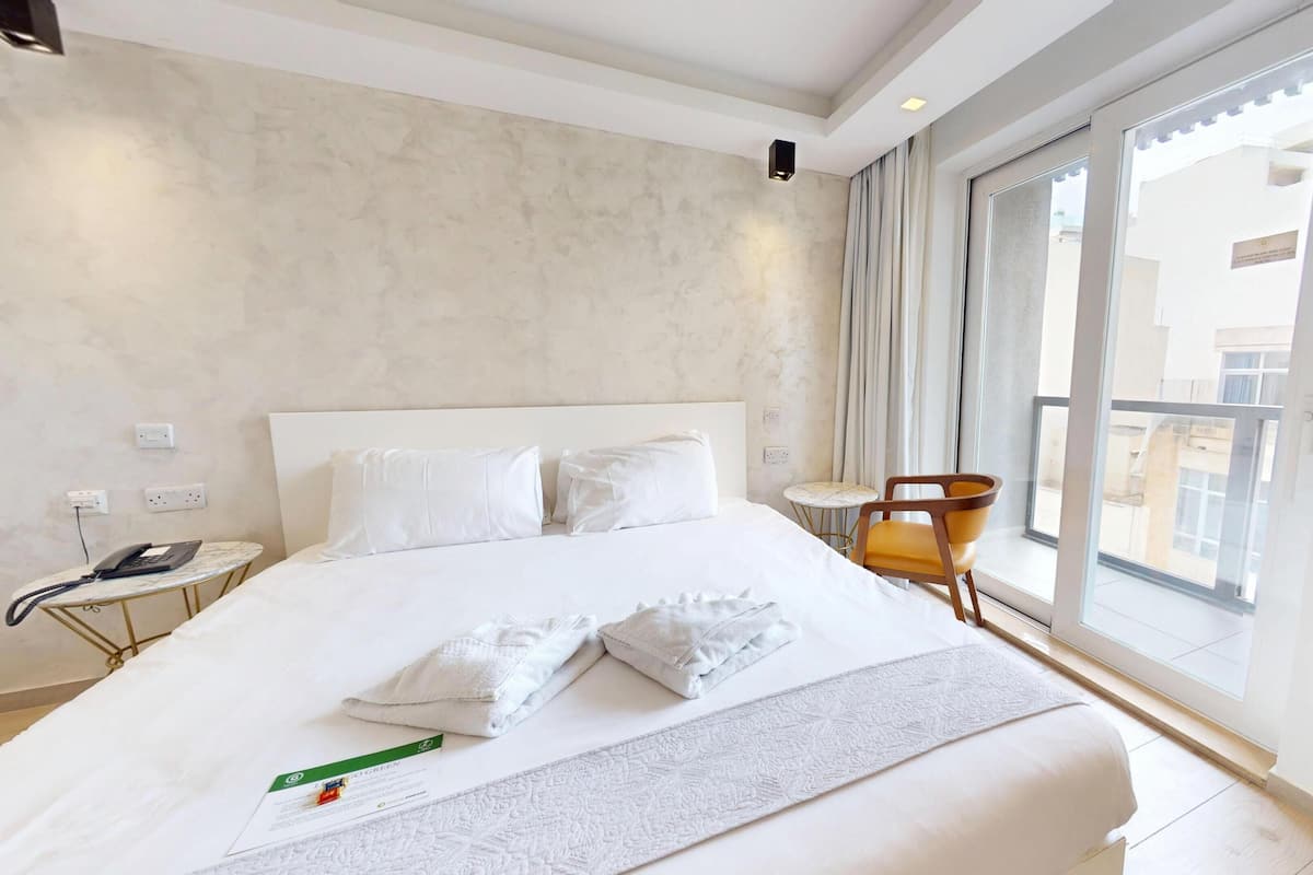 Quarto de hotel com uma cama grande, uma mesinha redonda, uma cadeira, janela com cortinas e vista para a varanda.