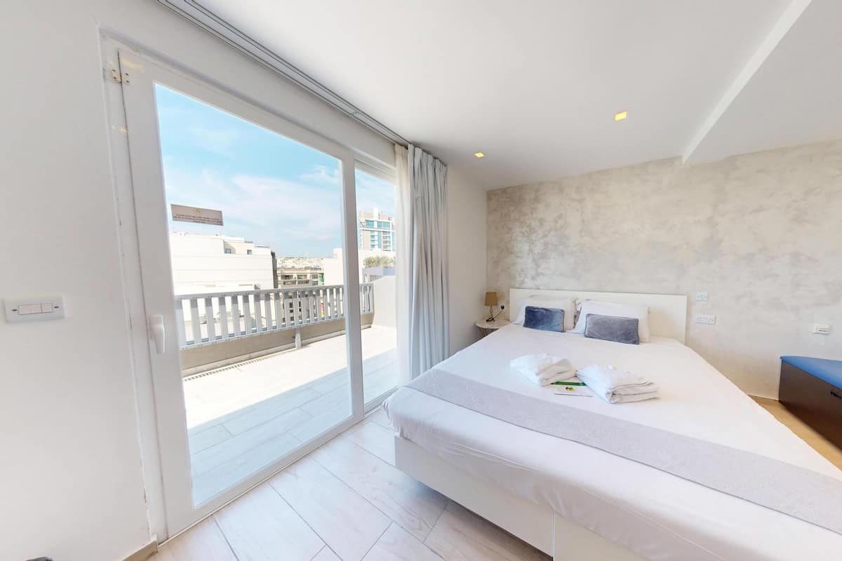 Quarto de hotel moderno com uma cama grande, uma escrivaninha, varanda com vista para edifícios e uma porta de vidro deslizante.
