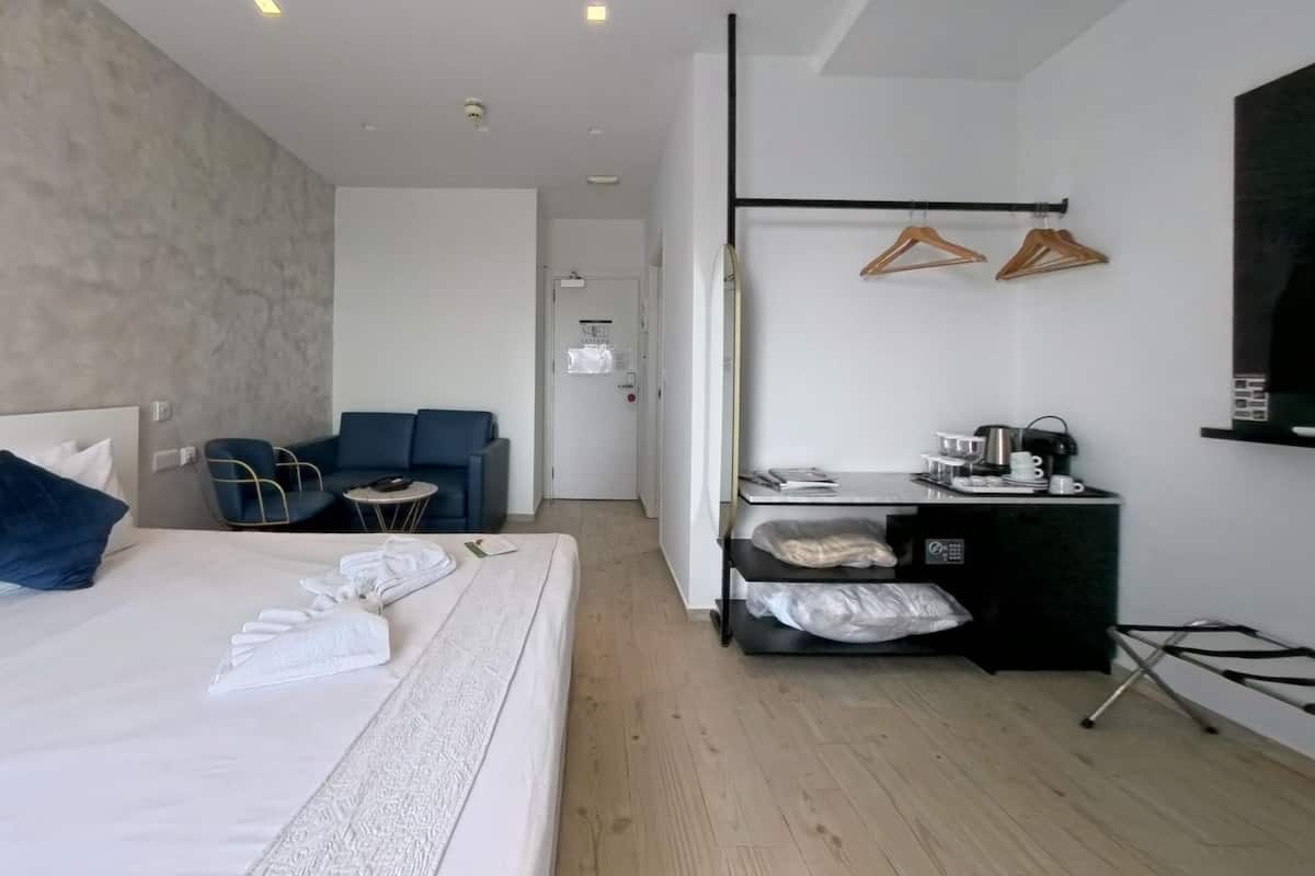 Quarto de hotel moderno com cama, área de estar com sofá e poltronas, mesa pequena, cozinha compacta e TV fixada na parede.