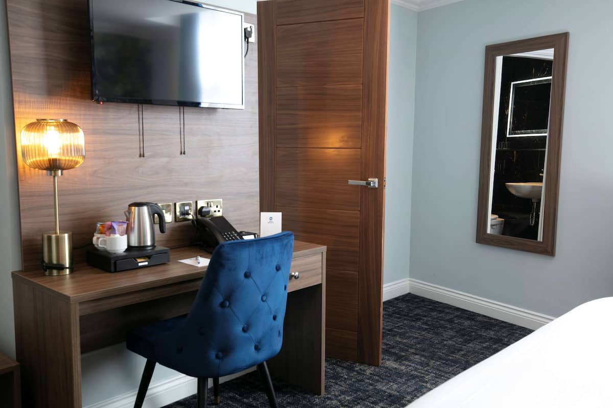 Quarto de hotel com escrivaninha de madeira, uma cadeira azul, televisão de tela plana na parede, mesa de cabeceira com abajur e um espelho grande.