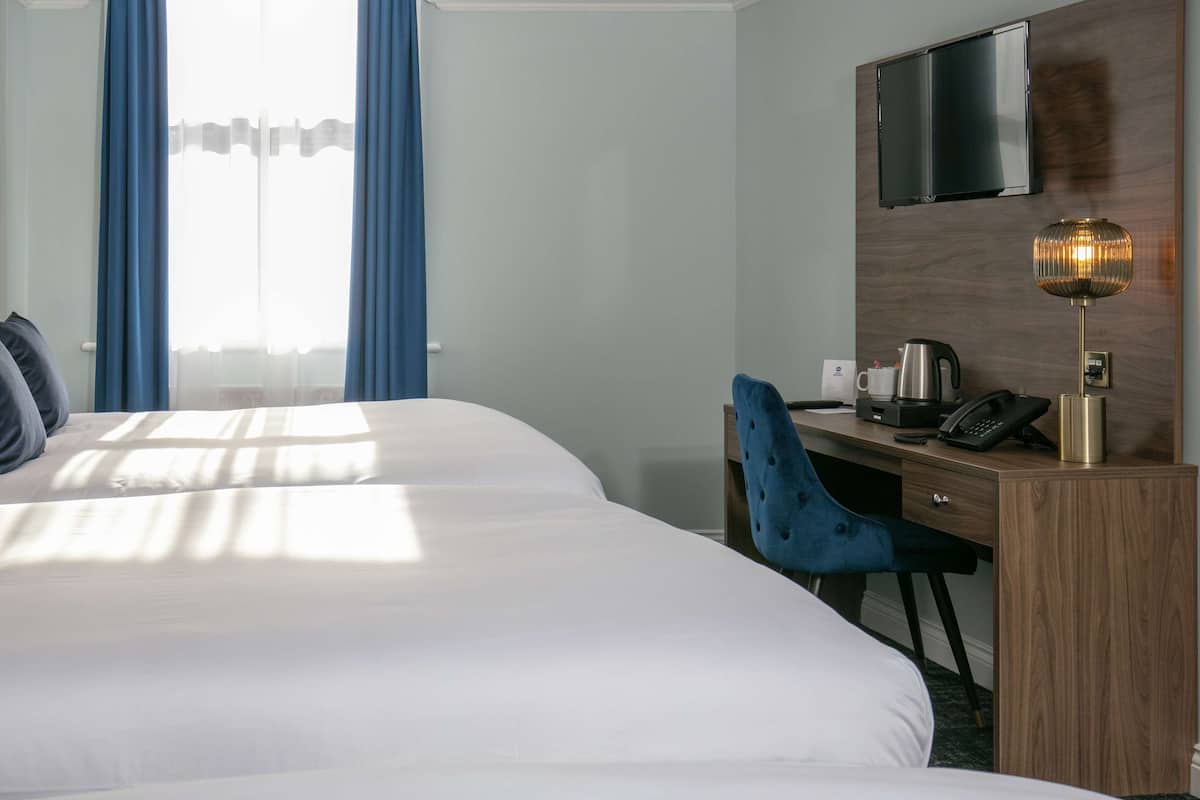 Quarto de hotel com uma cama grande, uma escrivaninha com luminária, uma cadeira, uma televisão e uma cafeteira.