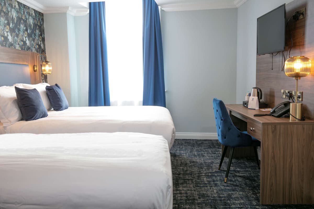 Quarto de hotel com duas camas, uma escrivaninha, uma cadeira, uma televisão e um abajur.