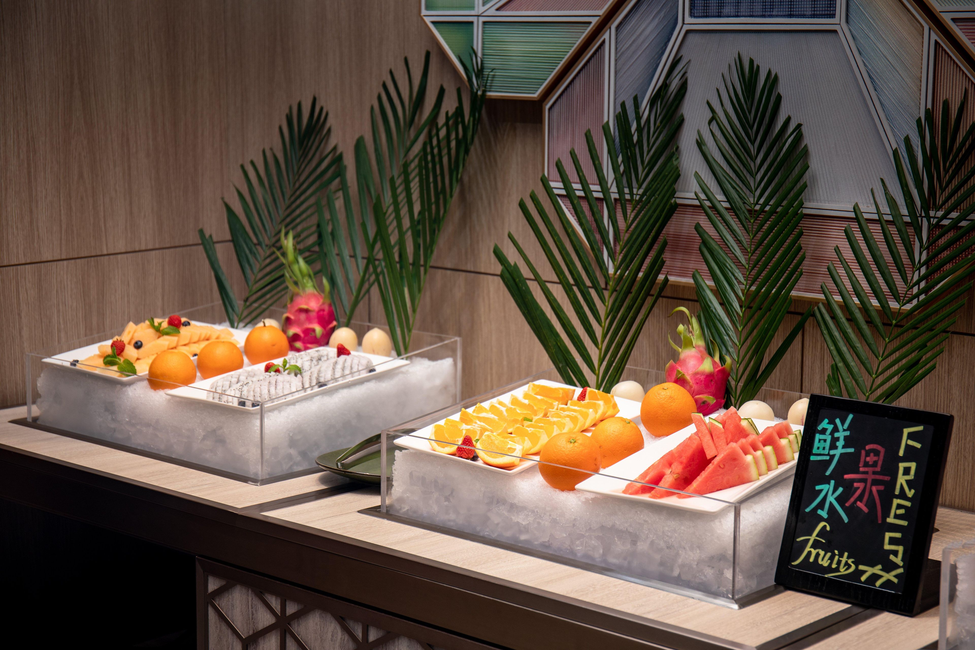 Exposição de frutas com gelo, plantas tropicais e um letreiro com nomes de frutas.