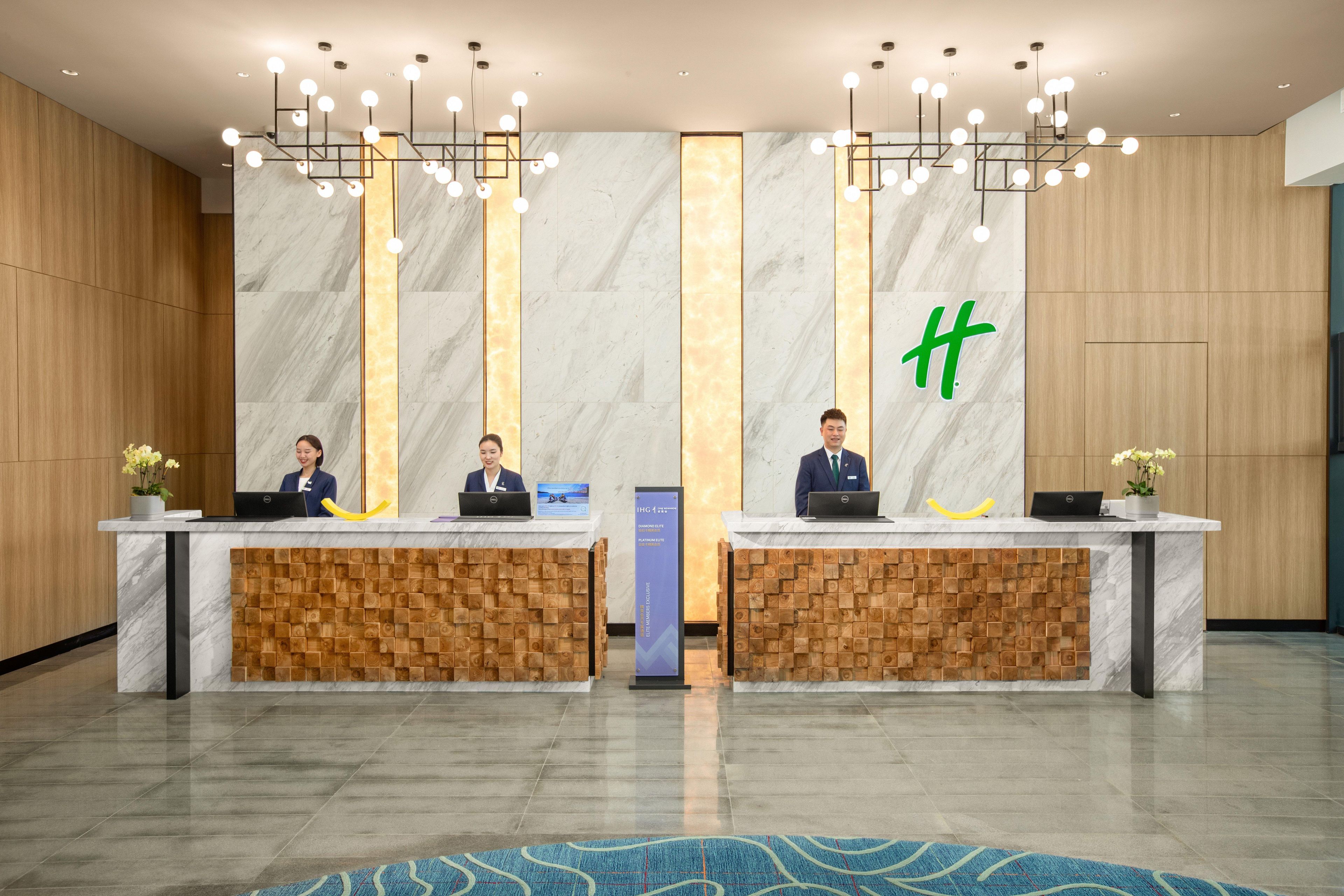 Bancada de recepção com três funcionários, parede de mármore e painel de madeira.