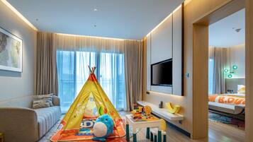 Quarto de hotel moderno com sofá, TV, cama, área de recreação com uma tenda e brinquedos, e uma janela grande com cortinas.