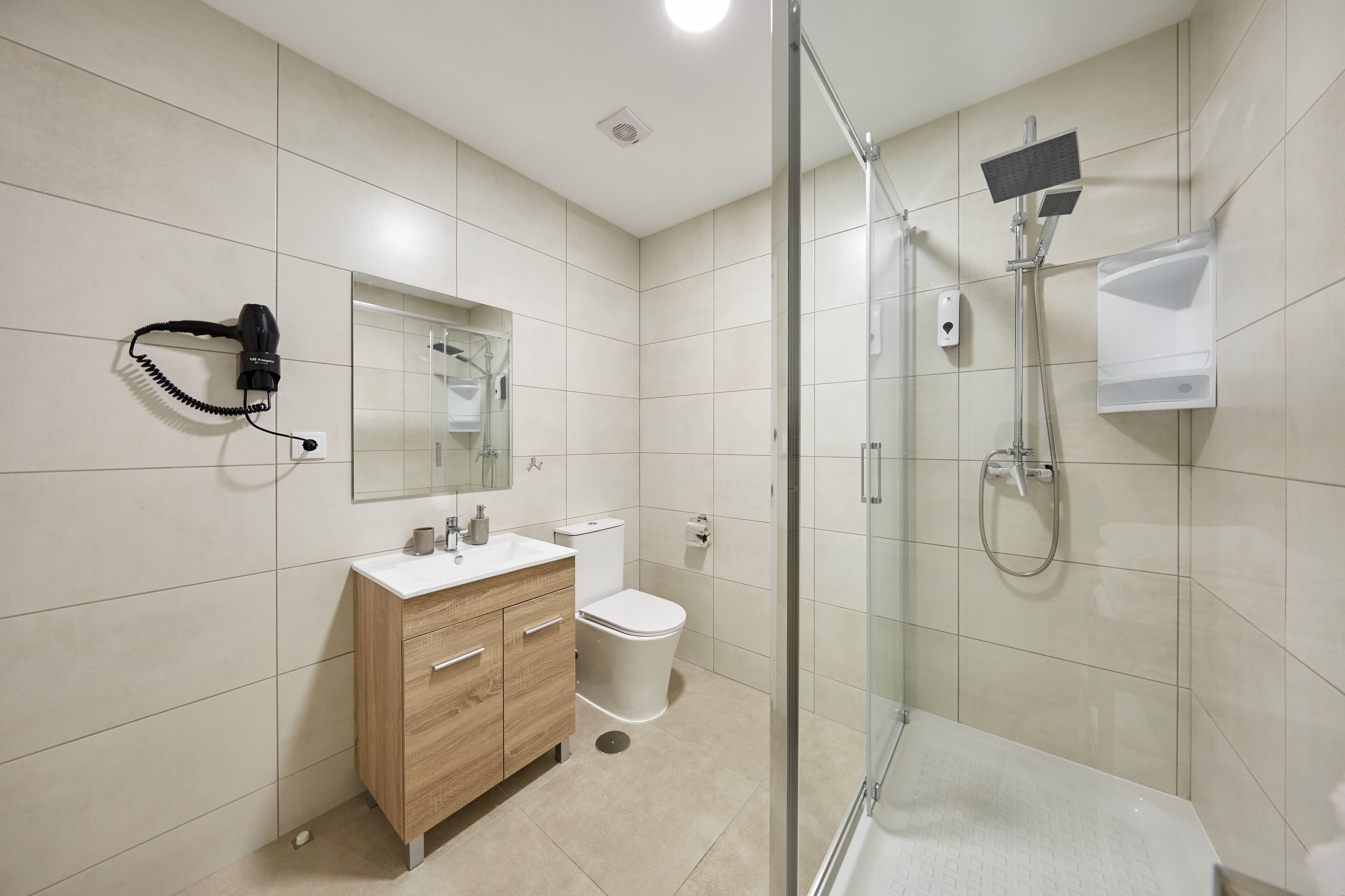 Uma casa de banho moderna com chuveiro em cabine de vidro, toucador de madeira com lavatório branco e secador de cabelo fixado na parede.
