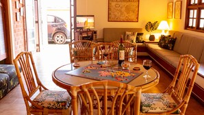 Uma área de refeições com uma mesa redonda, cadeiras e um jogo de tabuleiro. Há um sofá e um abajur ao fundo.