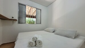 Quarto com cama, almofadas, janela com persianas e uma pequena prateleira com uma planta.