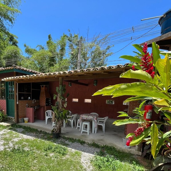 Uma área de pátio com muro de tijolos, um pequeno forno de barro, cadeiras de plástico branco e uma mesa com toalha de mesa florida.