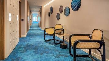 Corredor de hotel com carpete azul, uma fileira de cadeiras e obras de arte nas paredes.