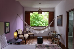 Quarto com rede, cadeiras, uma mesa pequena, cama e janela com vista para o verde.