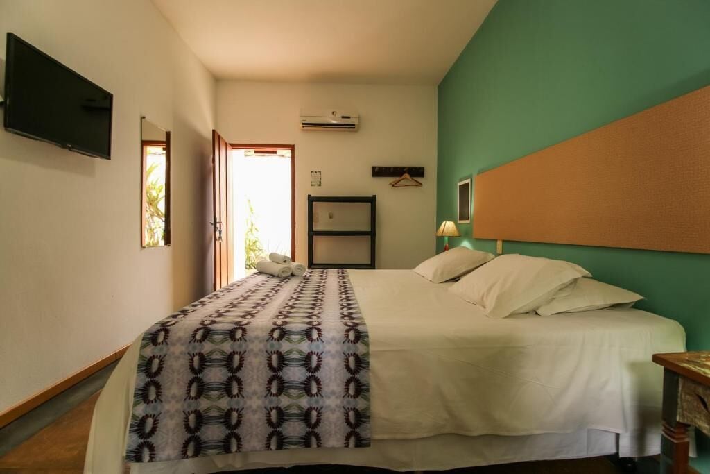 Quarto com uma cama, televisão na parede, ar condicionado e porta que dá para outro cômodo.
