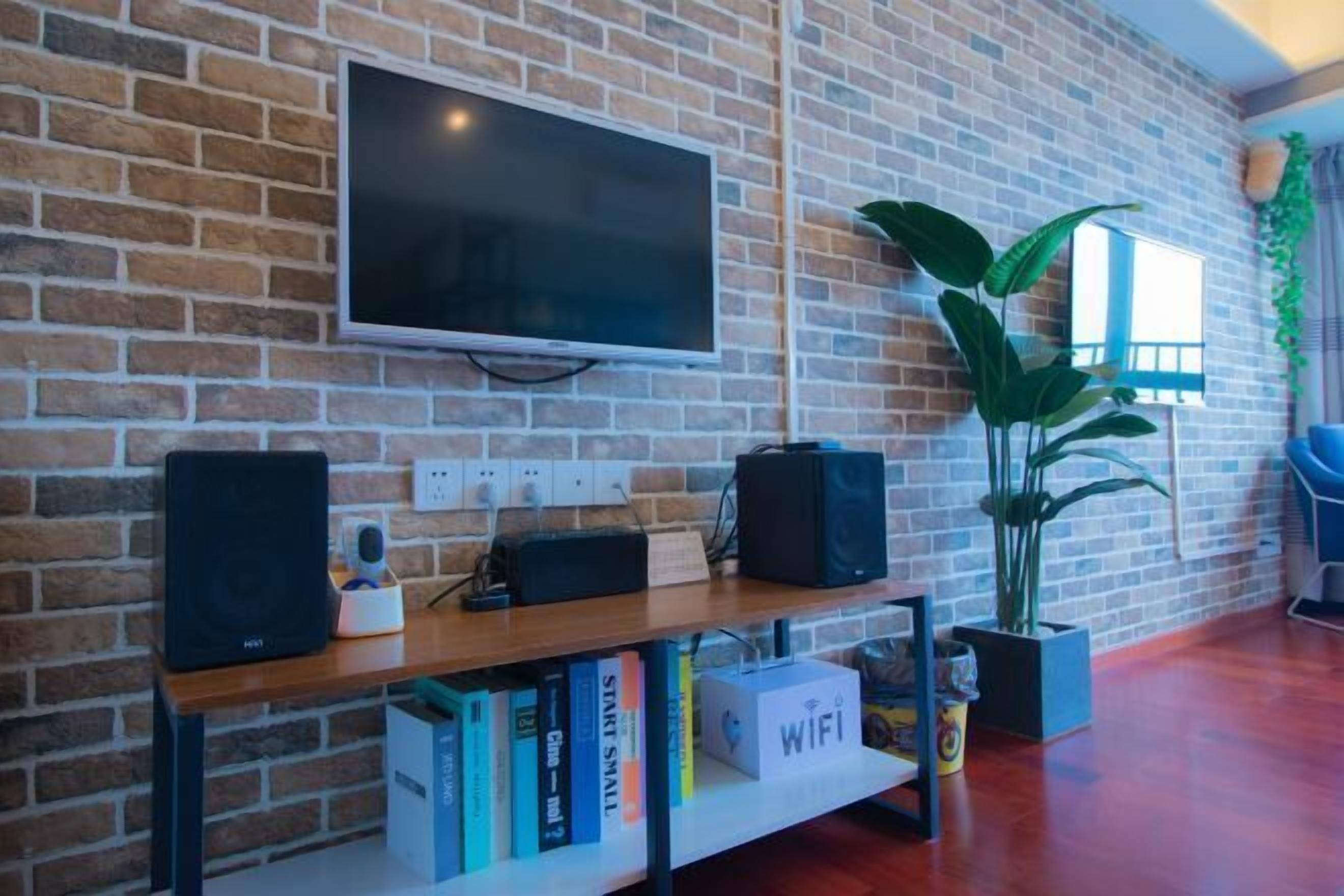 Quarto com parede de tijolos, televisão montada, prateleira de madeira com livros e uma placa 'WIFI', além de um vaso com planta.