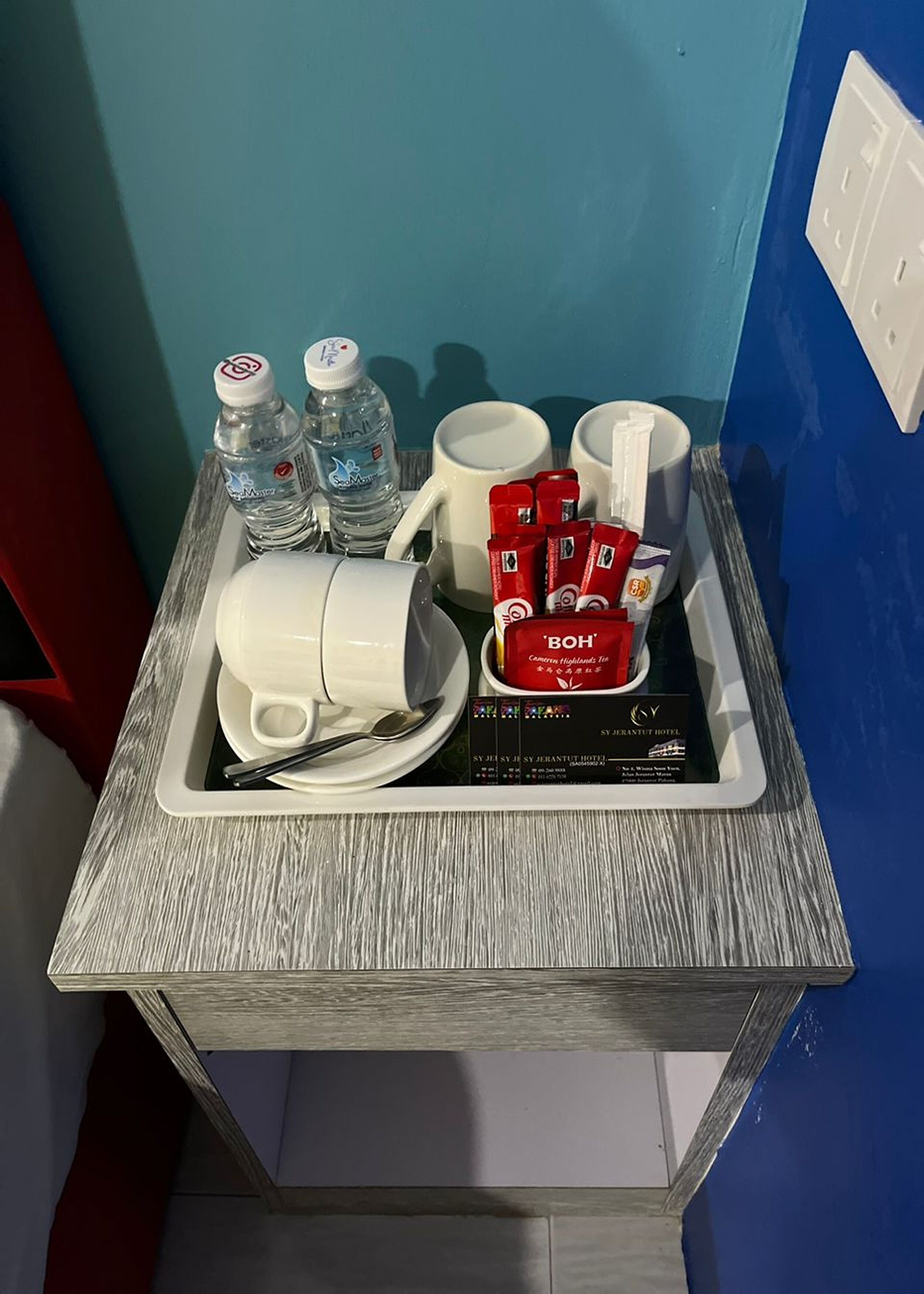 Uma bandeja com uma xícara, colher, duas garrafas de água e uma bandeja com sachês de chá vermelho em um ambiente de hotel.