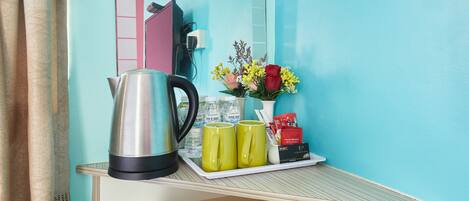 Uma área de cozinha compacta com chaleira, duas canecas, um suporte com sachês de café, um vaso com flores e um espelho refletindo um objeto rosa.