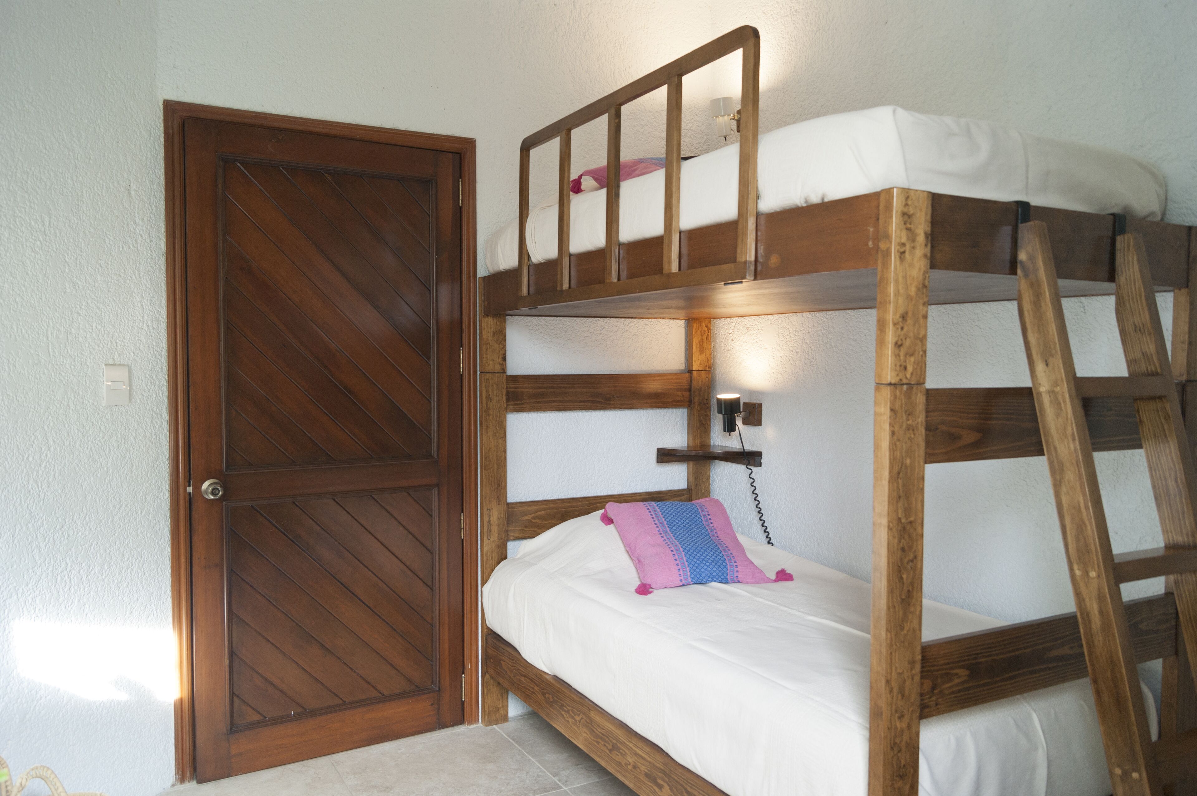 Uma cama beliche com estrutura de madeira, colchão branco e almofada listrada em rosa e azul. Há uma porta de madeira com padrão diagonal e uma pequena prateleira acima da cama.