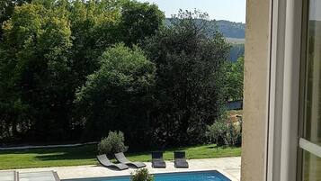 Vista de uma piscina com espreguiçadeiras, rodeada por vegetação e árvores, vista da janela.