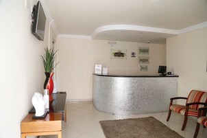 Uma área de recepção de hotel com balcão, televisão na parede, planta em vaso e uma cadeira listrada.