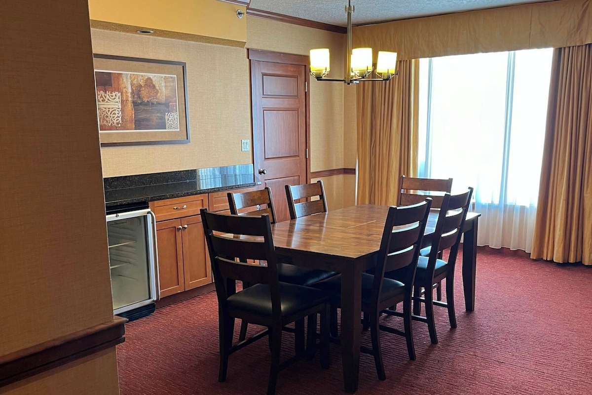Uma sala de jantar com mesa e cadeiras de madeira, área de cozinha com geladeira e armários, e uma grande janela com cortinas.