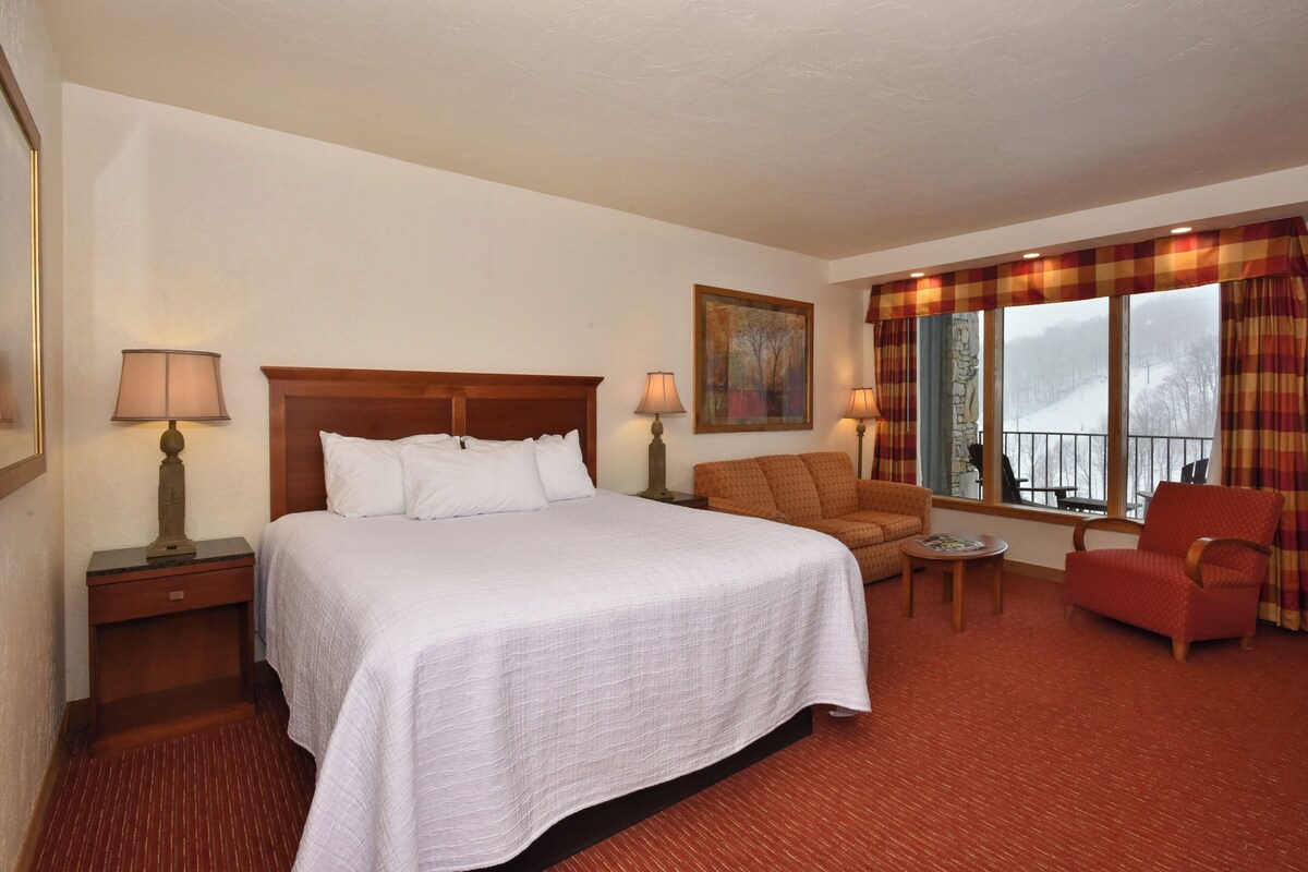 Quarto de hotel com uma cama grande, duas mesas de cabeceira, uma cadeira, uma mesinha e uma janela com cortinas.