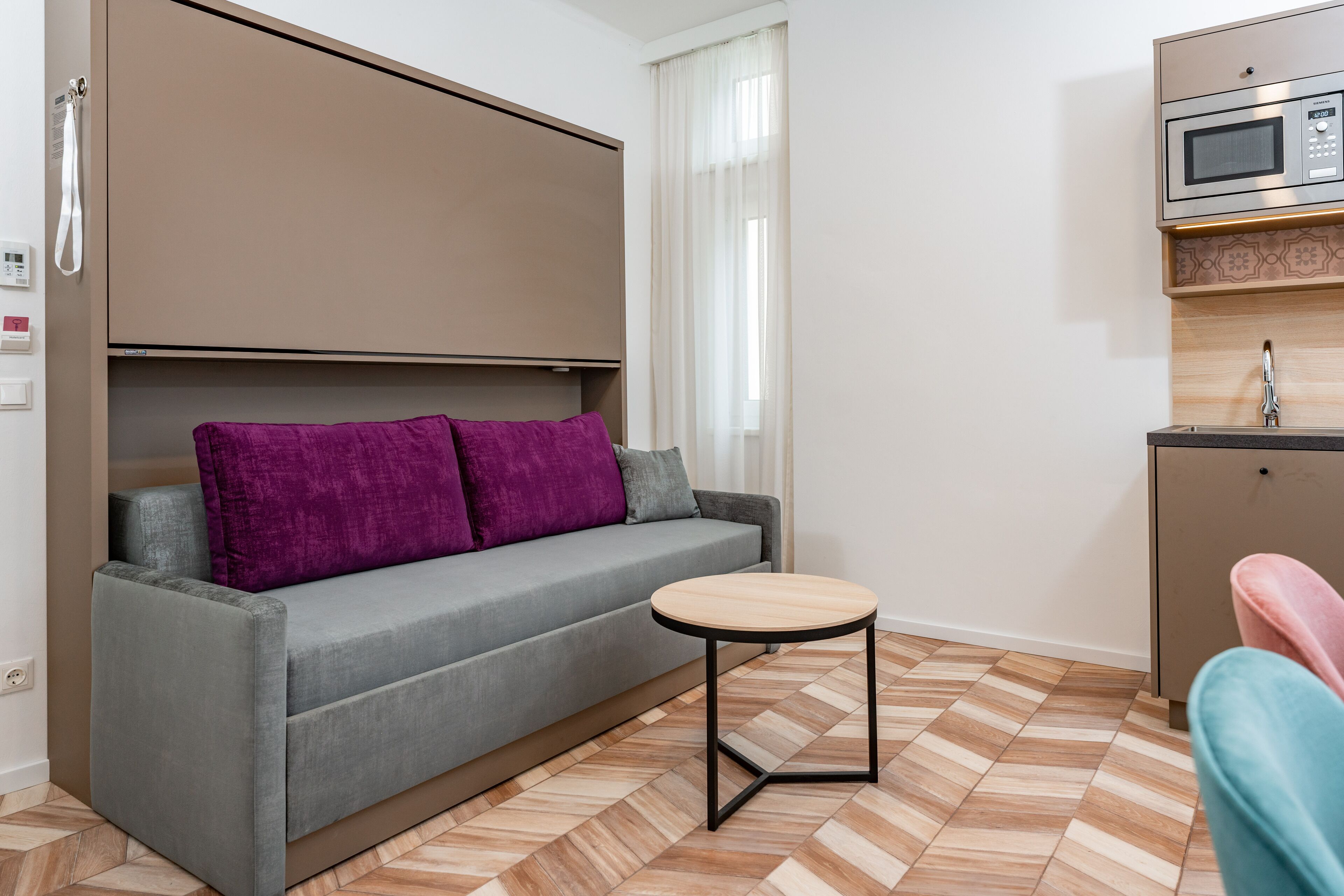 Um espaço de estar compacto com sofá, mesa redonda pequena, cozinha compacta e janela com cortinas.