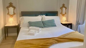 Cama bem arrumada com lençóis brancos e manta amarela mostarda, ladeada por duas mesas de cabeceira com luminárias, em um quarto com design simples e clean.