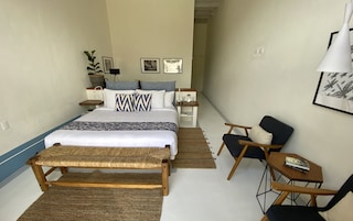 Quarto de hotel com cama, um banco, uma cadeira, uma mesinha e uma mesa de cabeceira.