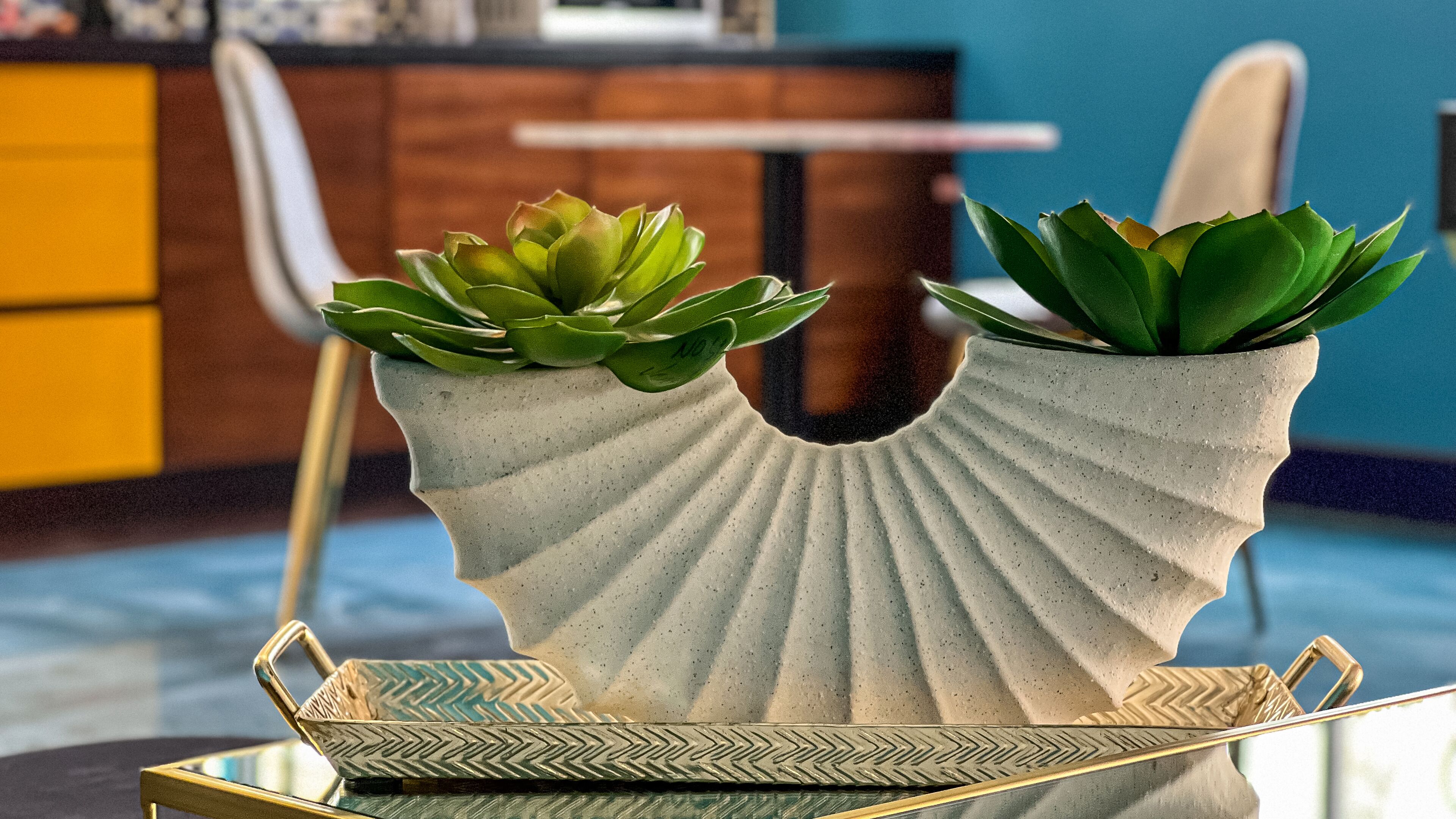 Suporte decorativo para plantas com duas suculentas, um suporte estampado e uma cadeira num quarto com parede azul e um armário de madeira.