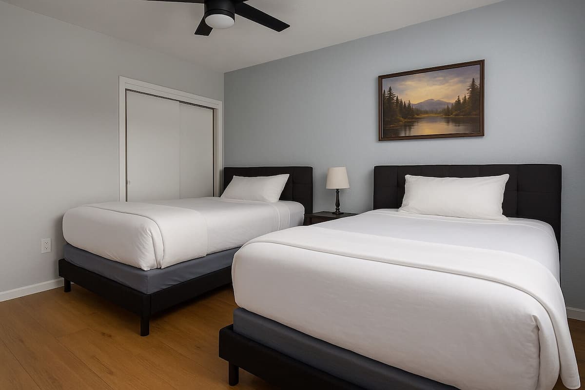 Quarto de hotel com duas camas, um ventilador de teto, um quadro de paisagem e uma porta.