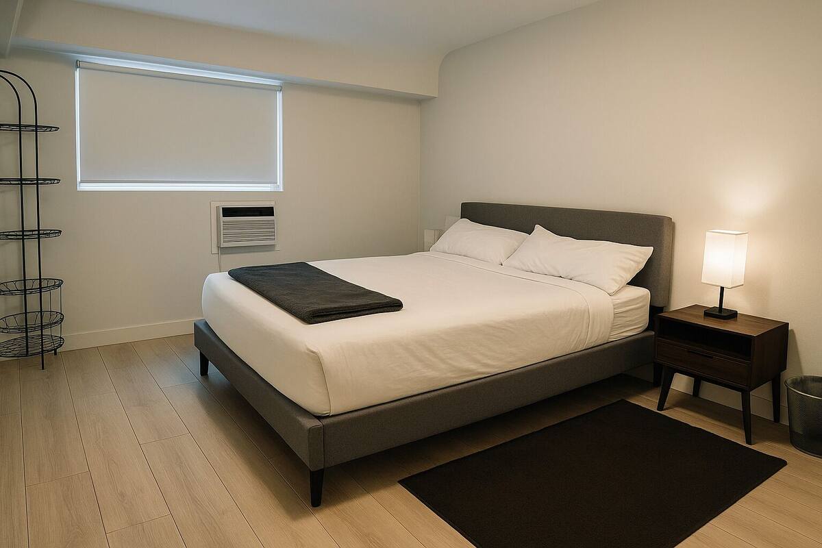 Cama bem arrumada com cabeceira cinza, luminária de cabeceira e janela com persianas.