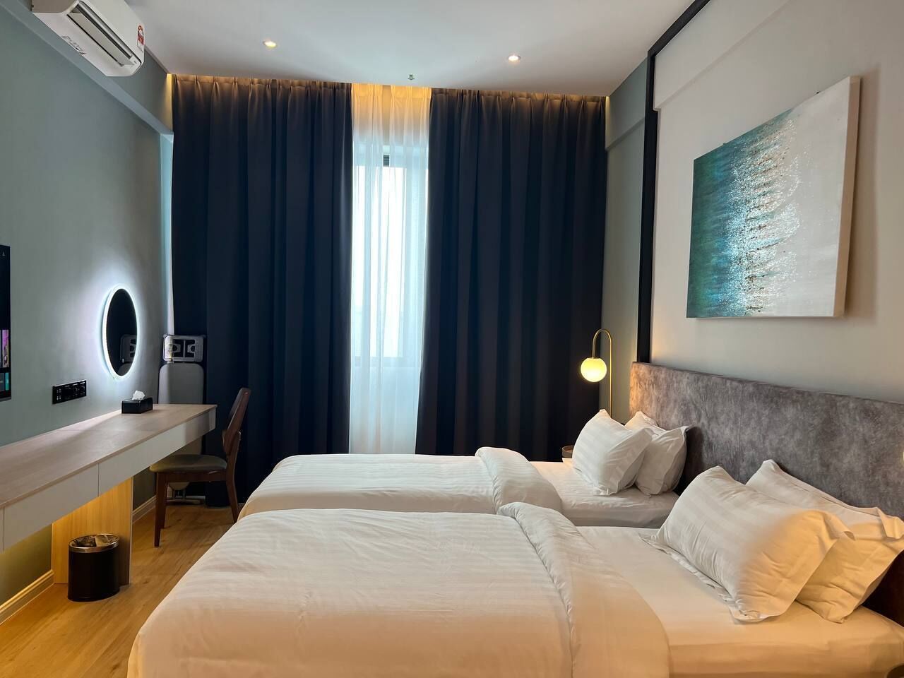 Quarto de hotel com duas camas, uma escrivaninha com computador, televisão e uma grande janela com cortinas.