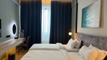 Quarto de hotel com duas camas, uma escrivaninha com computador, televisão e uma grande janela com cortinas.