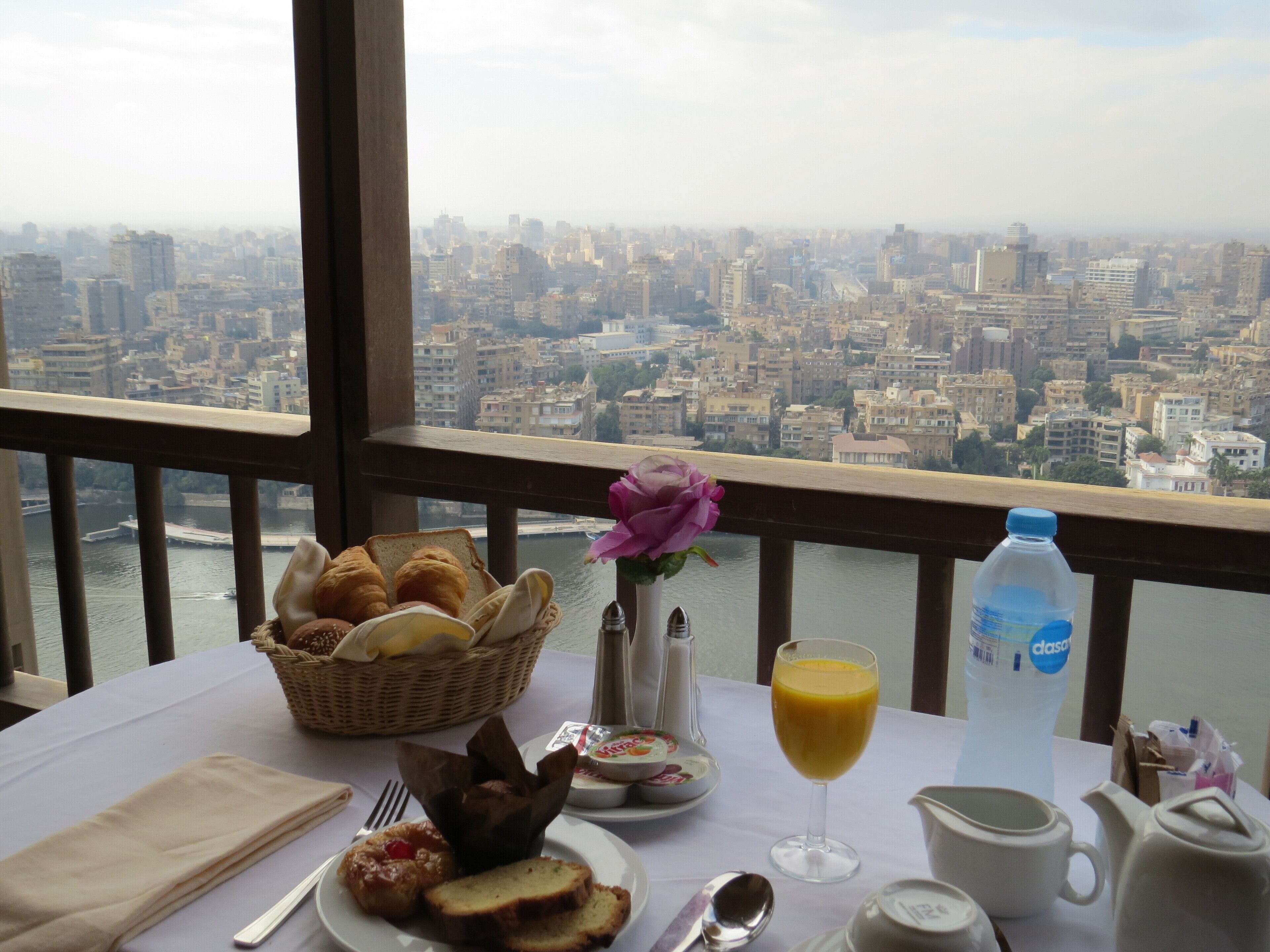 Mesa de café da manhã com croissants, um copo de suco de laranja e uma garrafa de água Dasani, com vista para um horizonte urbano com rio e edifícios.
