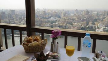 Mesa de café da manhã com croissants, um copo de suco de laranja e uma garrafa de água Dasani, com vista para um horizonte urbano com rio e edifícios.