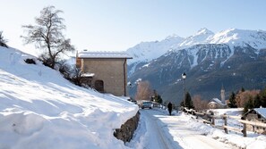 Uma estrada de montanha coberta de neve, com um edifício de pedra, uma cerca de madeira e o topo de uma igreja ao fundo.