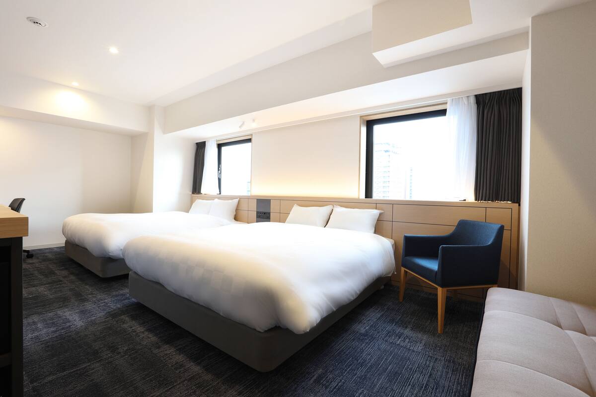 Quarto de hotel com duas camas, uma escrivaninha, uma cadeira e uma janela grande com cortinas.