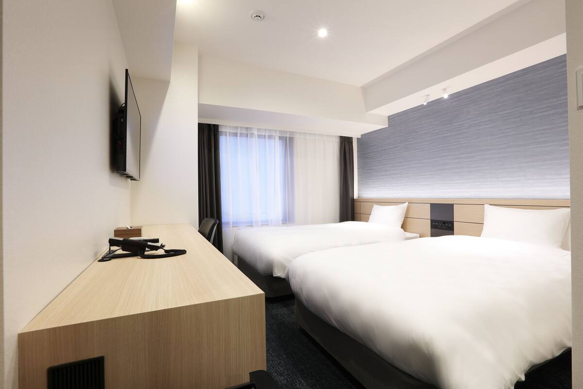 Quarto de hotel com cama, escrivaninha, televisão na parede e janela com cortinas.