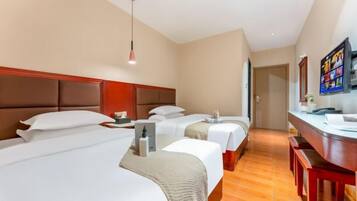 Quarto de hotel com três camas, cabeceira de madeira, escrivaninha e uma cadeira, e uma televisão.