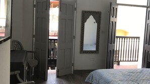 Quarto com cama, uma cadeira, guarda-roupa e espelho. Duas portas abertas dão para uma varanda.