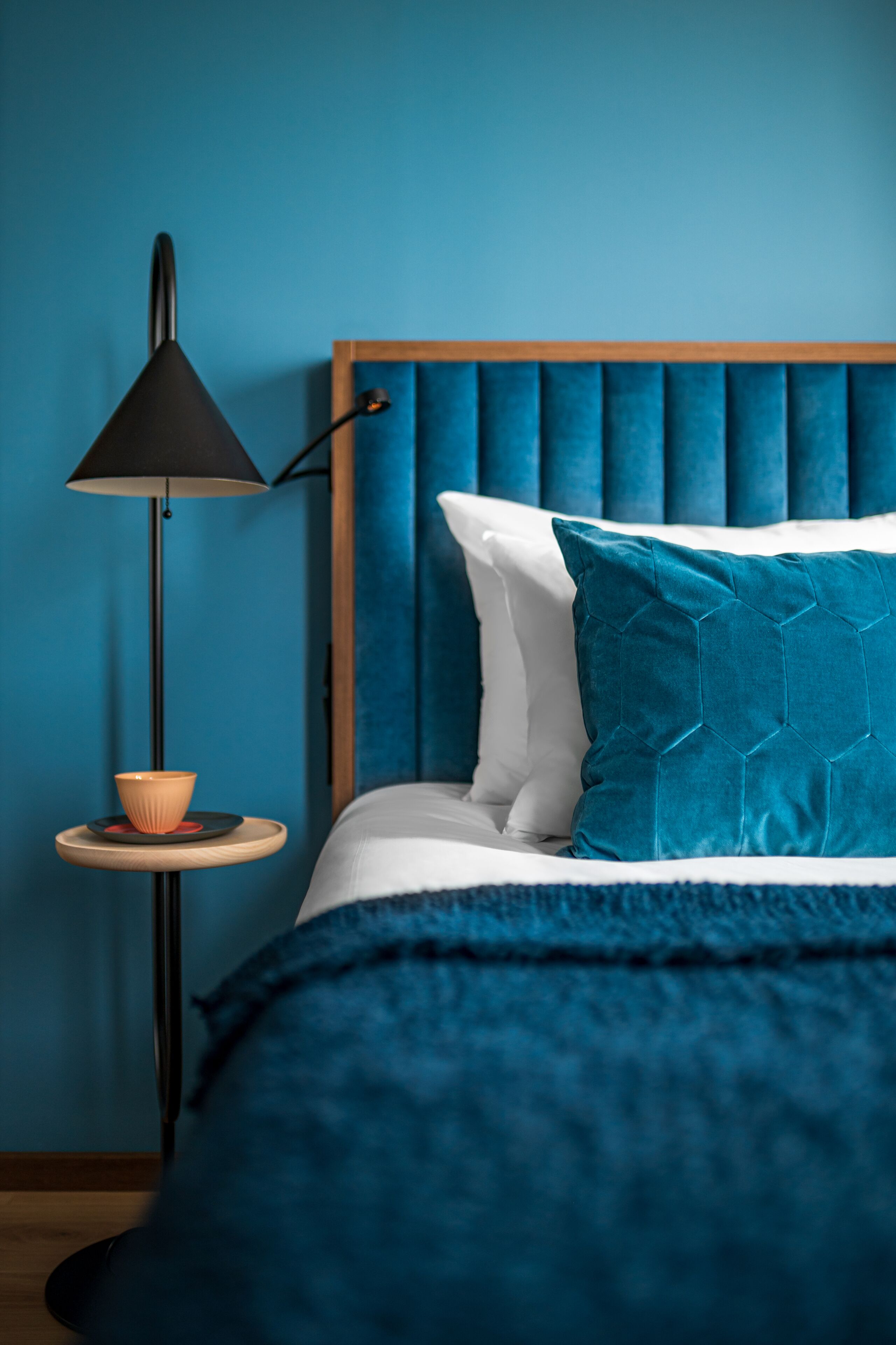 Quarto com parede azul, cabeceira de madeira, cama com colcha azul e branca, abajur de chão preto e mesa redonda pequena com uma tigela.
